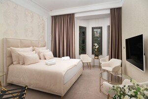Quarto de hotel com uma cama grande, duas cadeiras, uma mesinha, televisão e janela com cortinas.
