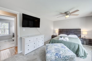 Quarto com cama, cômoda branca, ventilador de teto, TV fixada na parede e mesa de cabeceira com abajur.