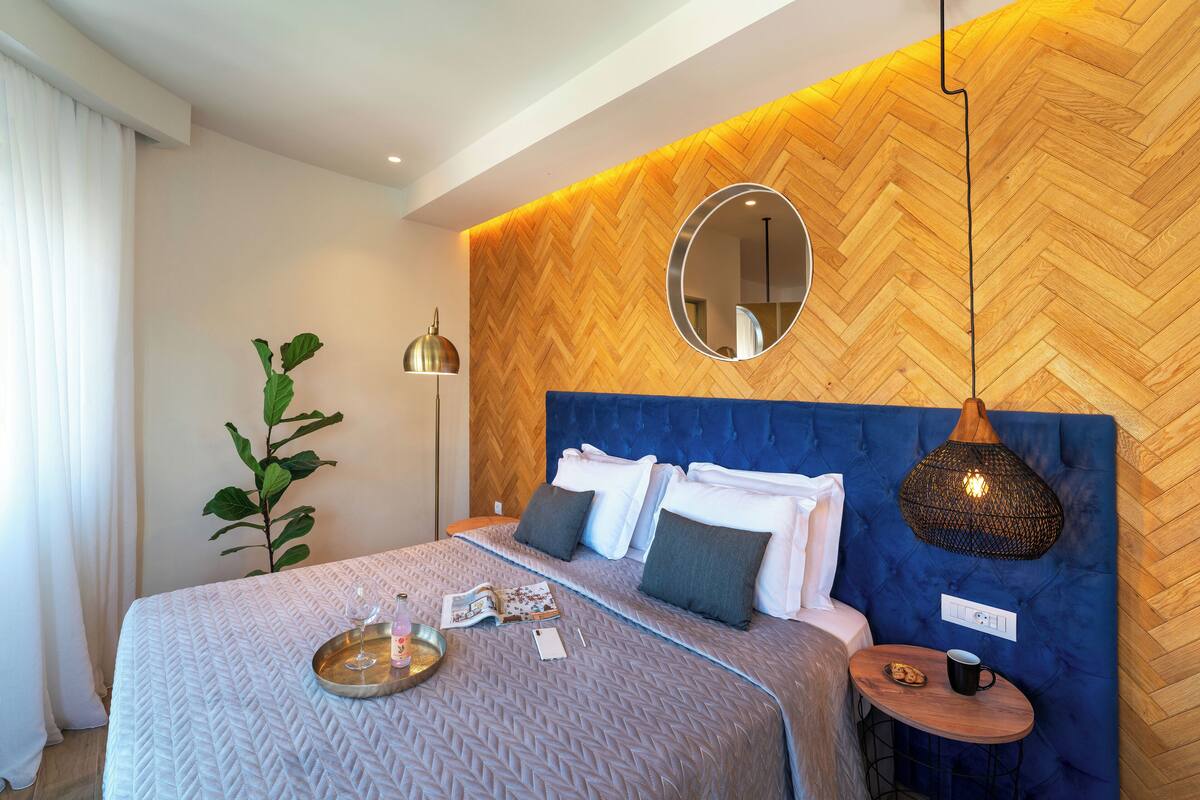 Quarto de hotel moderno com parede de madeira em padrão herringbone, cabeceira azul com efeito de matelassê e espelho redondo.