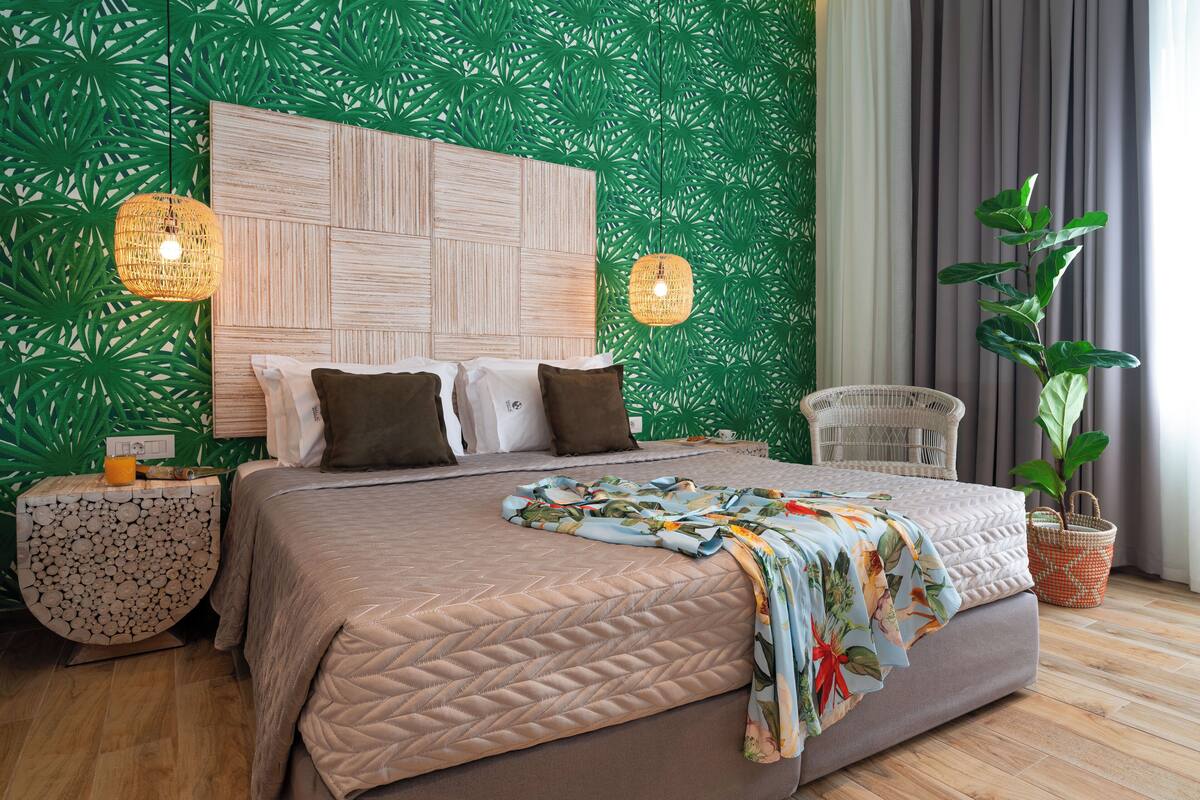 Quarto moderno com cabeceira de madeira, parede estampada, cama grande com colcha acolchoada, duas luminárias de cabeceira, uma poltrona de vime e uma planta em vaso.