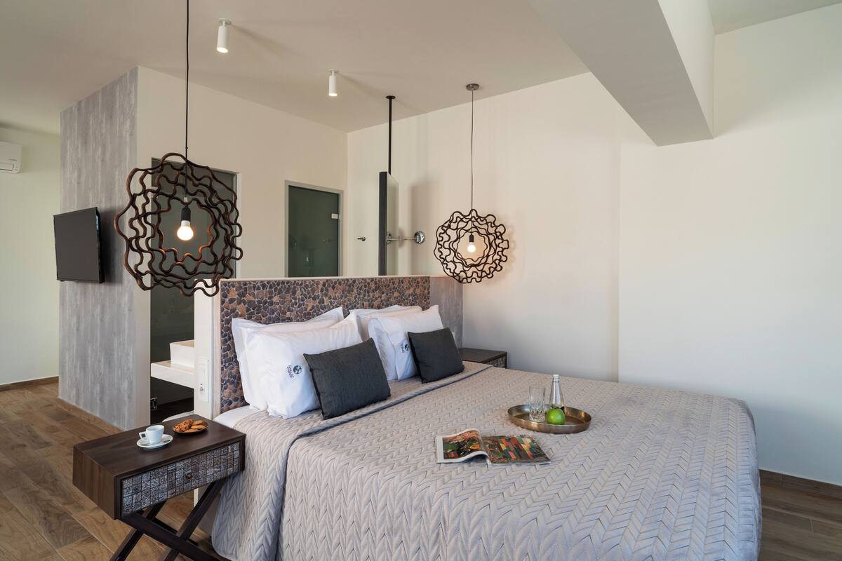 Quarto moderno com uma cama grande, mesa de cabeceira com uma chávena e um prato, uma revista e um objeto decorativo na parede.