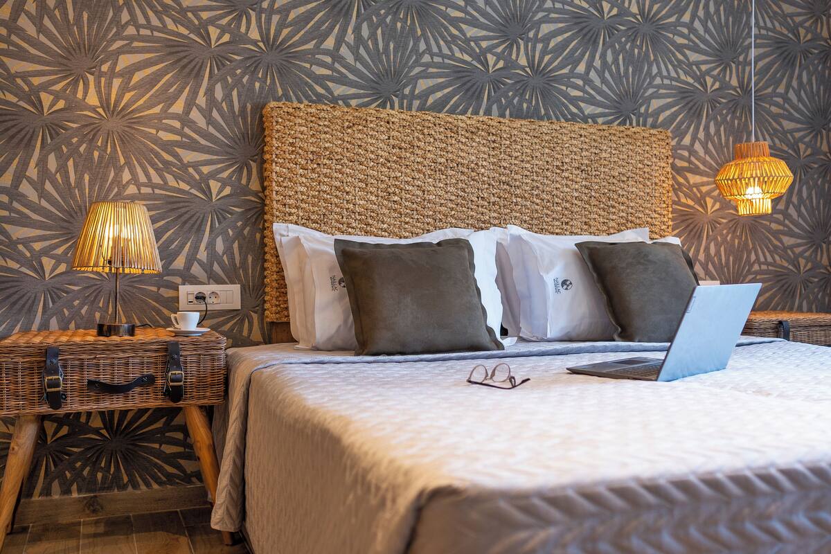 Cama bem arrumada com um laptop e óculos em cima, mesa de cabeceira de vime com uma luminária e uma xícara, e uma parede estampada.