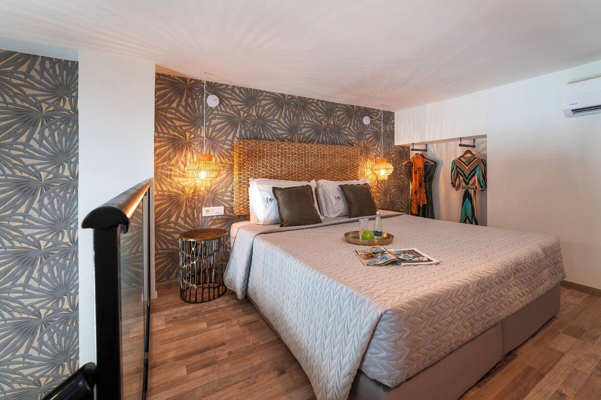 Quarto de hotel com uma cama grande, uma mesinha de cabeceira, parede com padrão geométrico e um cabideiro com roupas penduradas.