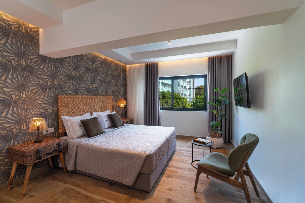 Um quarto moderno com uma cama grande, mesas de cabeceira, uma cadeira, uma janela com cortinas e uma TV fixada na parede.