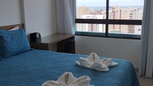 Quarto de hotel com uma cama grande, uma escrivaninha e vista para a cidade através de uma janela ampla.