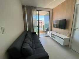 Uma sala de estar moderna com um sofá preto, uma TV montada na parede e uma porta de vidro deslizante que dá para uma varanda.