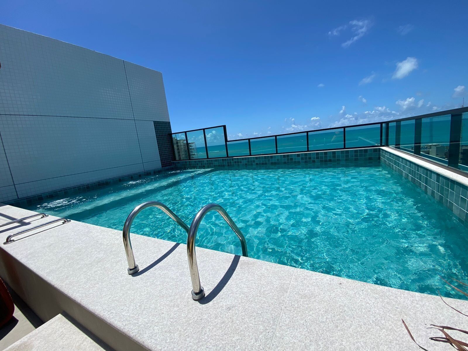 Uma piscina azul límpida com grades de metal, rodeada por um muro de azulejos brancos e uma barreira de vidro com vista para o oceano.