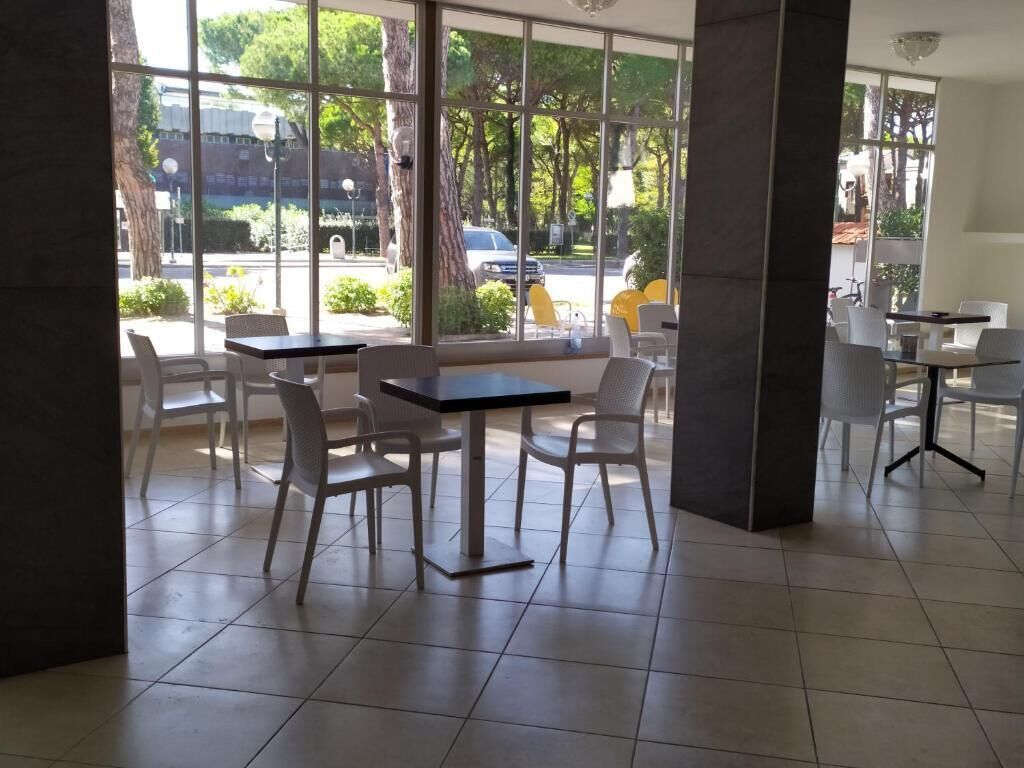 Amplo espaço interno com grandes janelas, cadeiras e mesas brancas dispostas para refeições.