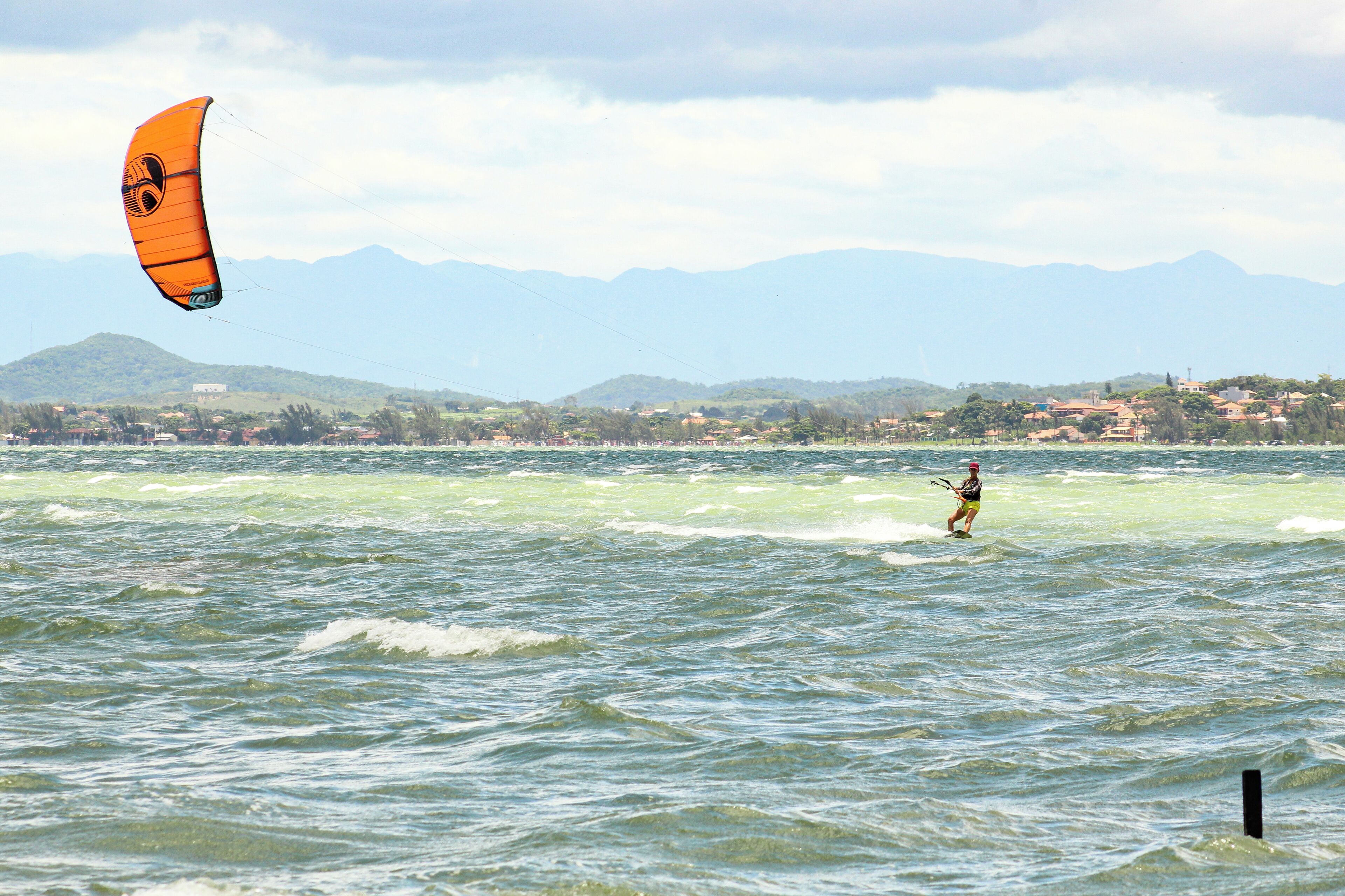 Uma pessoa praticando windsurf num lago, com montanhas ao fundo.