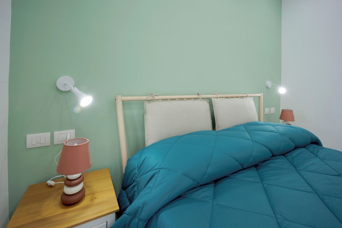 Quarto com colcha azul-turquesa, mesa de cabeceira de madeira com abajur e luminária na parede.