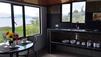 Cozinha moderna com uma janela ampla para um lago, área de jantar com mesa posta para dois e bancada com utensílios de cozinha.