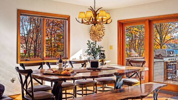 Uma sala de jantar com uma grande mesa de madeira, cadeiras, um lustre e vista para árvores de outono através de amplas janelas.