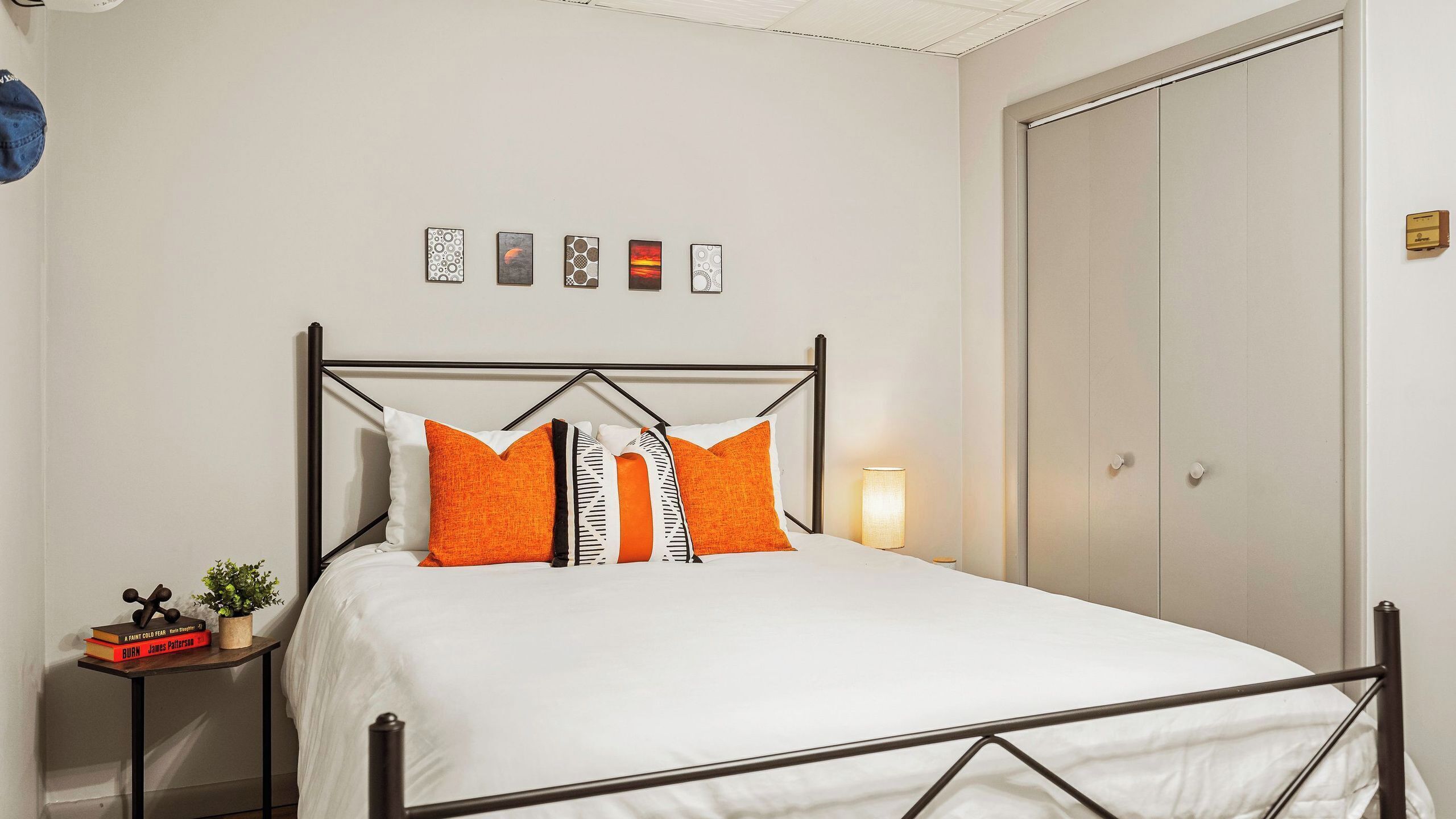 Cama com estrutura metálica, roupa de cama branca e almofadas laranja. Há quadros nas paredes acima da cama e uma porta à direita.