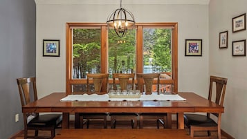 Uma sala de jantar com uma grande mesa de madeira, seis cadeiras, um candelabro e janelas amplas com vista para árvores e um corpo d'água.
