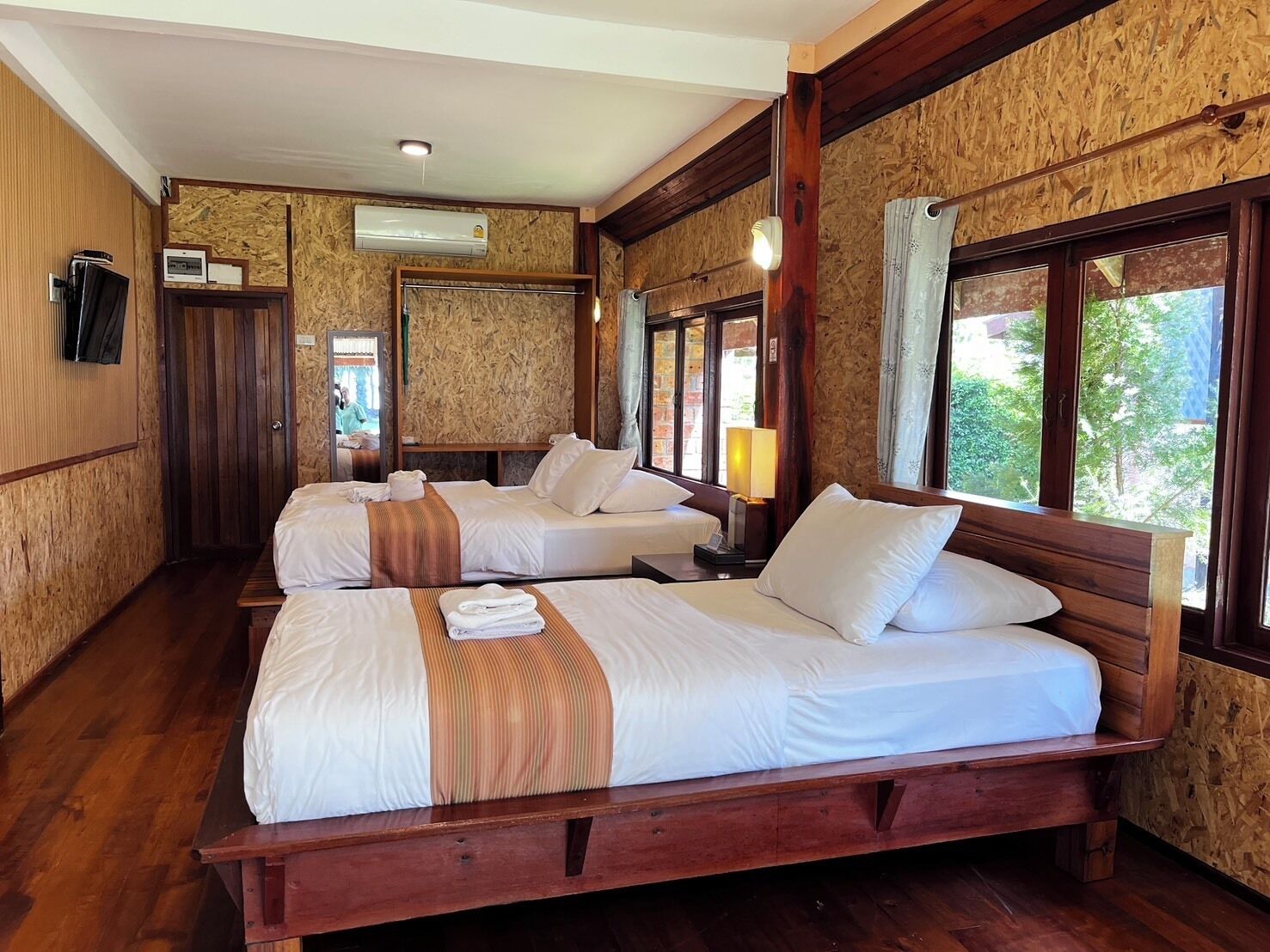 Quarto de hotel com duas camas, piso de madeira, uma janela grande com vista e um aparelho de ar condicionado na parede.