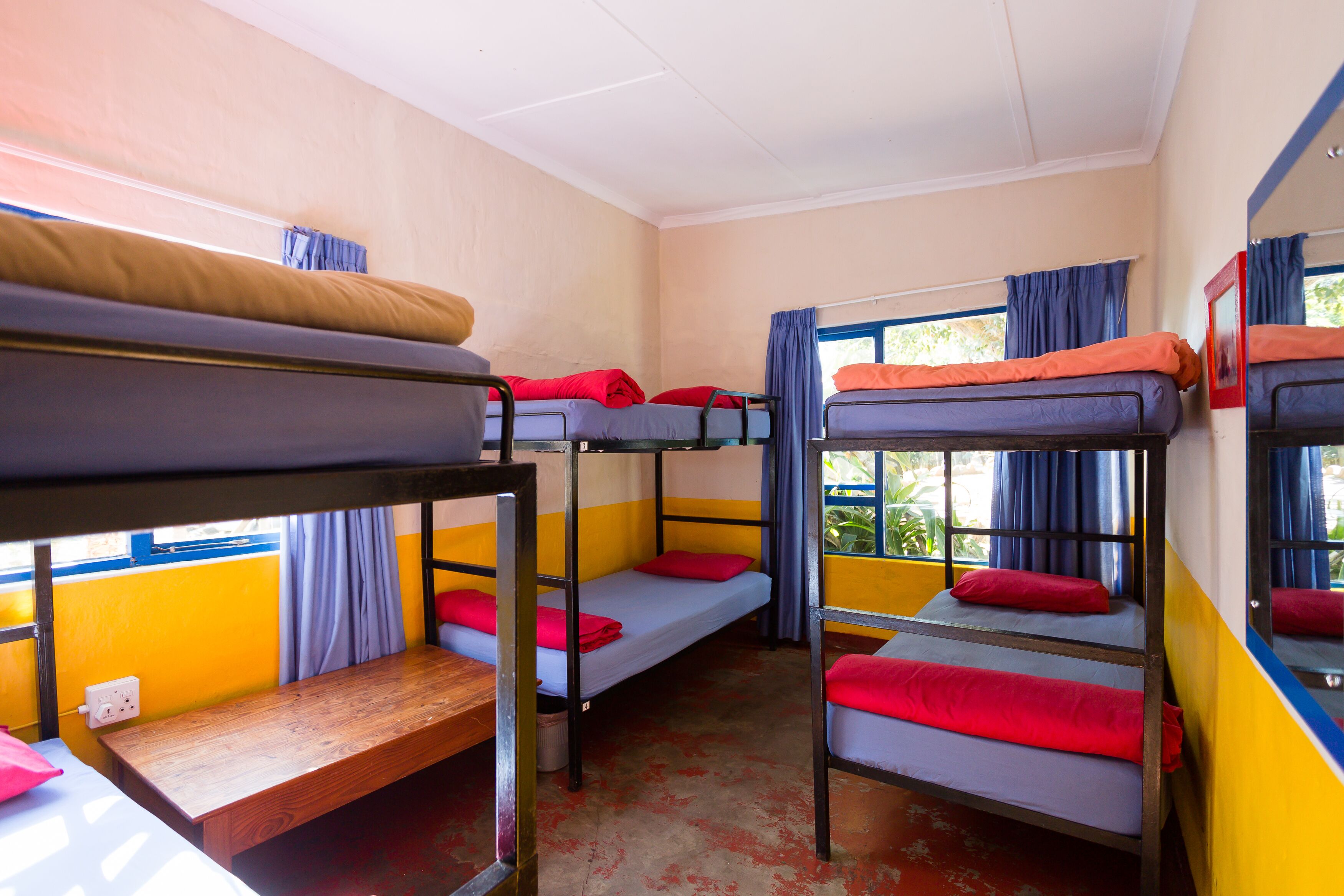 Um quarto de estudante com beliches, uma escrivaninha de madeira e uma janela com cortinas.