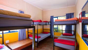 Um quarto de estudante com beliches, uma escrivaninha de madeira e uma janela com cortinas.
