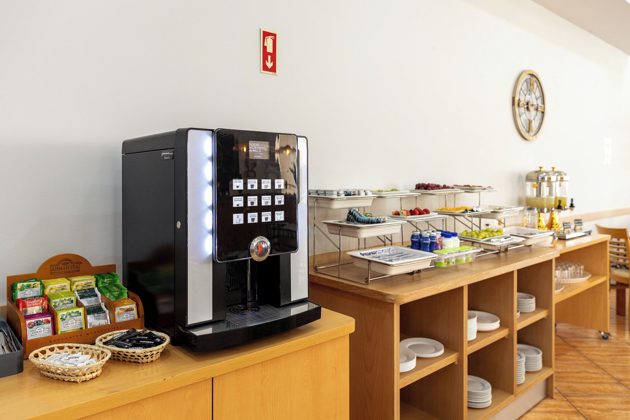 Máquina de café com diversas opções, dispensador de chá e área de buffet com vários alimentos e acompanhamentos.