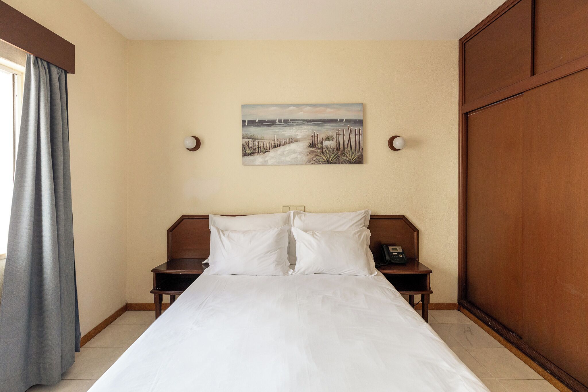 Quarto de hotel com uma cama, uma mesinha de cabeceira, um quadro na parede e um guarda-roupa de madeira.