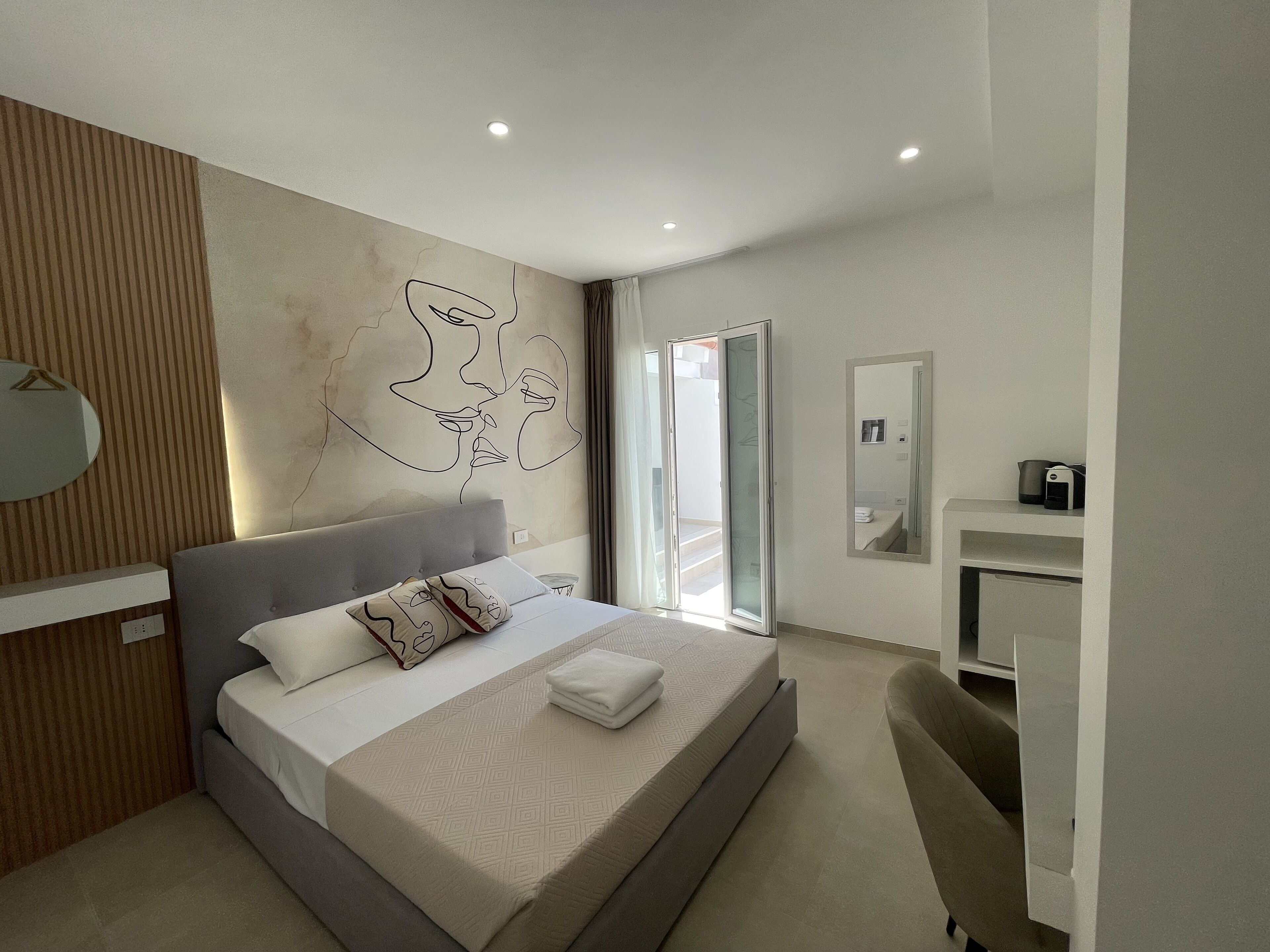Quarto moderno com uma cama grande, uma mesa de cabeceira, uma cadeira, uma escrivaninha com espelho e uma obra de arte na parede.