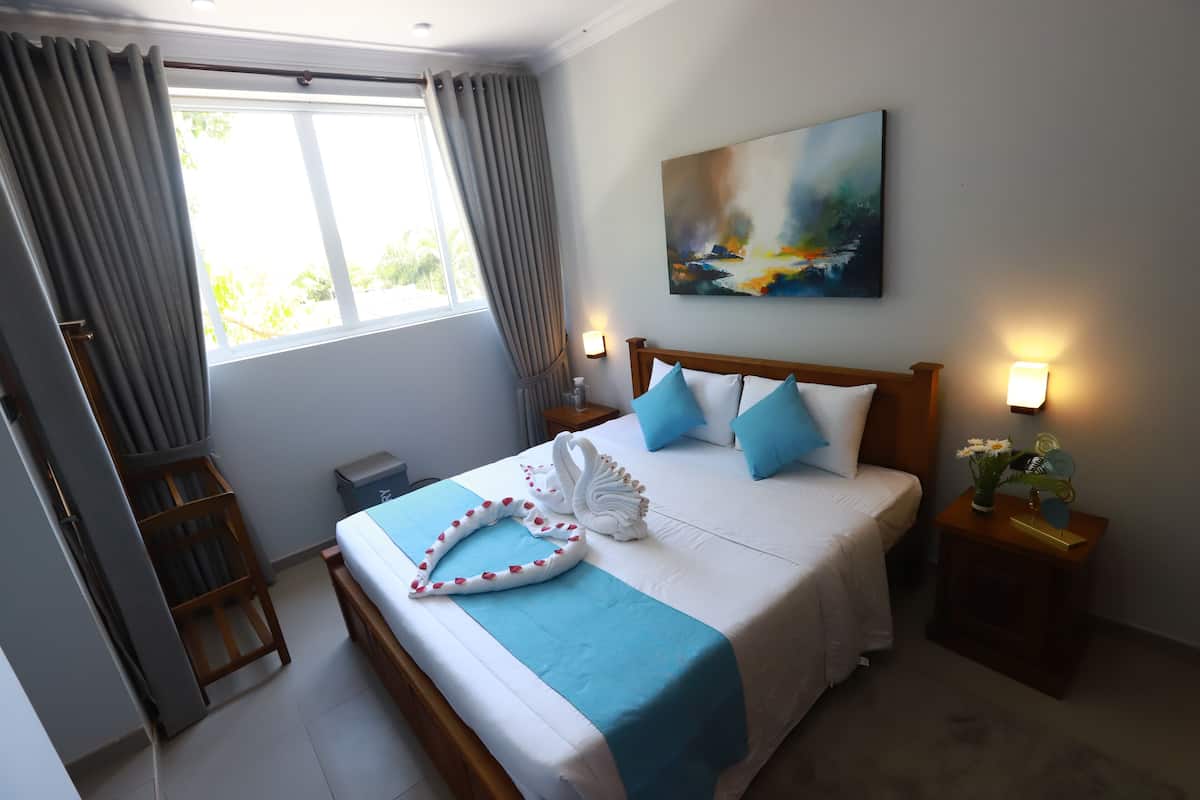 Cama bem arrumada com colcha azul, travesseiros brancos e toalha em forma de cisne branco. Há um quadro na parede, uma janela com cortinas e uma mesa de cabeceira com abajur e um vaso de flores.