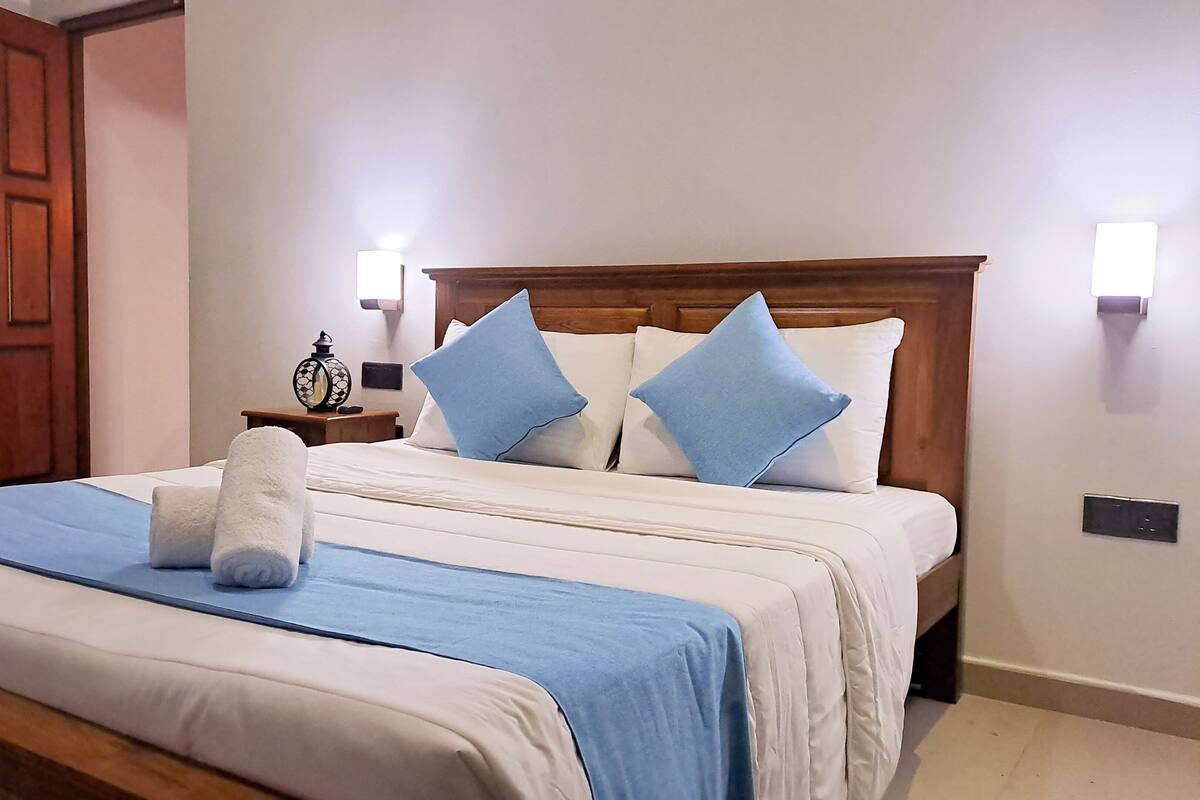 Cama bem arrumada com roupa de cama azul e branca, cabeceira de madeira e duas luminárias de cabeceira.