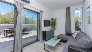 Uma sala de estar moderna com um sofá, uma mesa de centro e uma televisão. Há uma porta de vidro deslizante que dá para uma área externa com uma mesa e cadeiras.