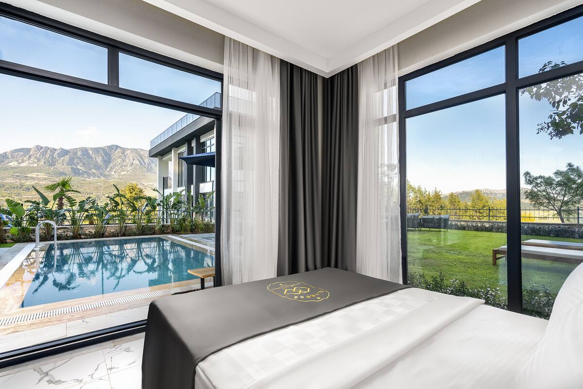 Quarto de hotel moderno com uma grande janela, uma cama e vista para uma piscina e montanhas.