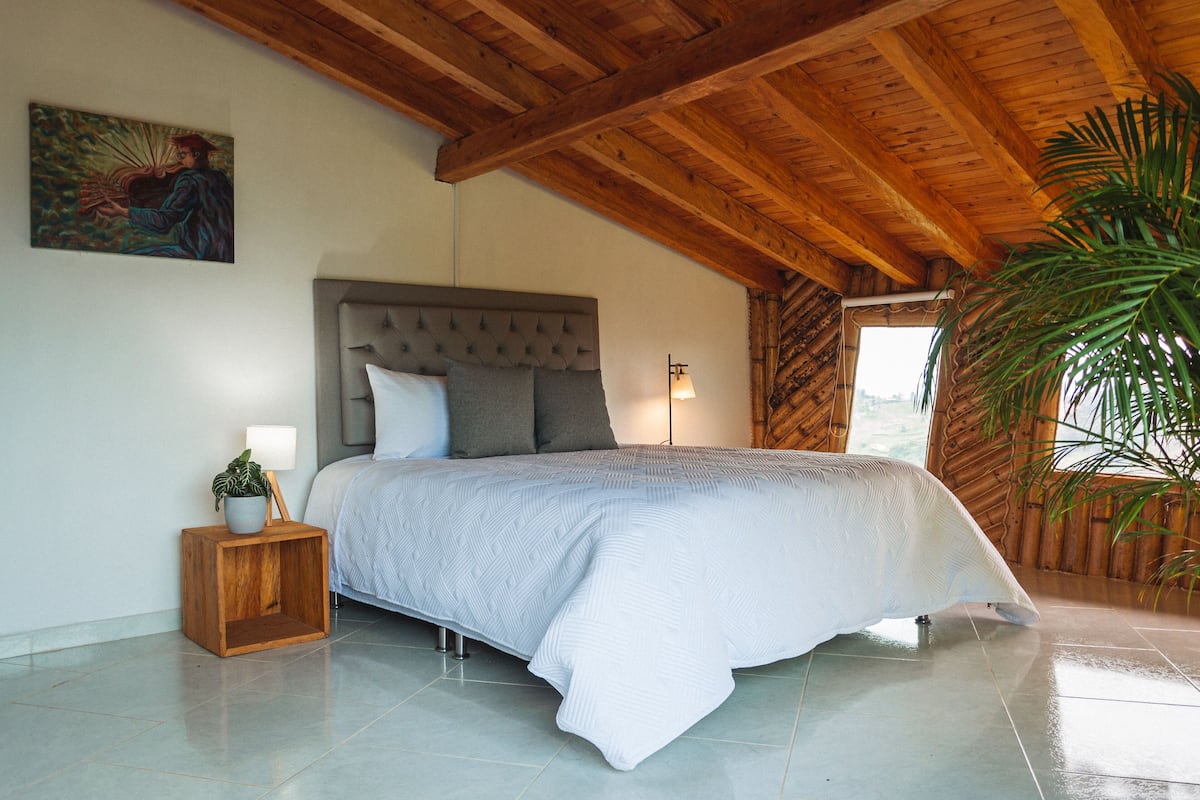 Quarto com teto de madeira, cama com cabeceira cinza, mesa de cabeceira com luminária e um vaso de planta.