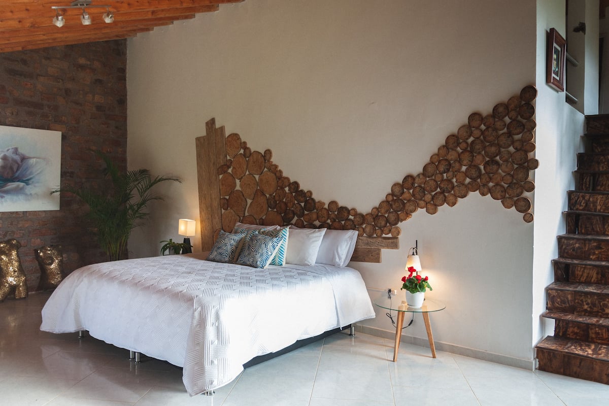 Quarto com uma cama grande, cabeceira de madeira, uma mesinha com abajur e uma escada.