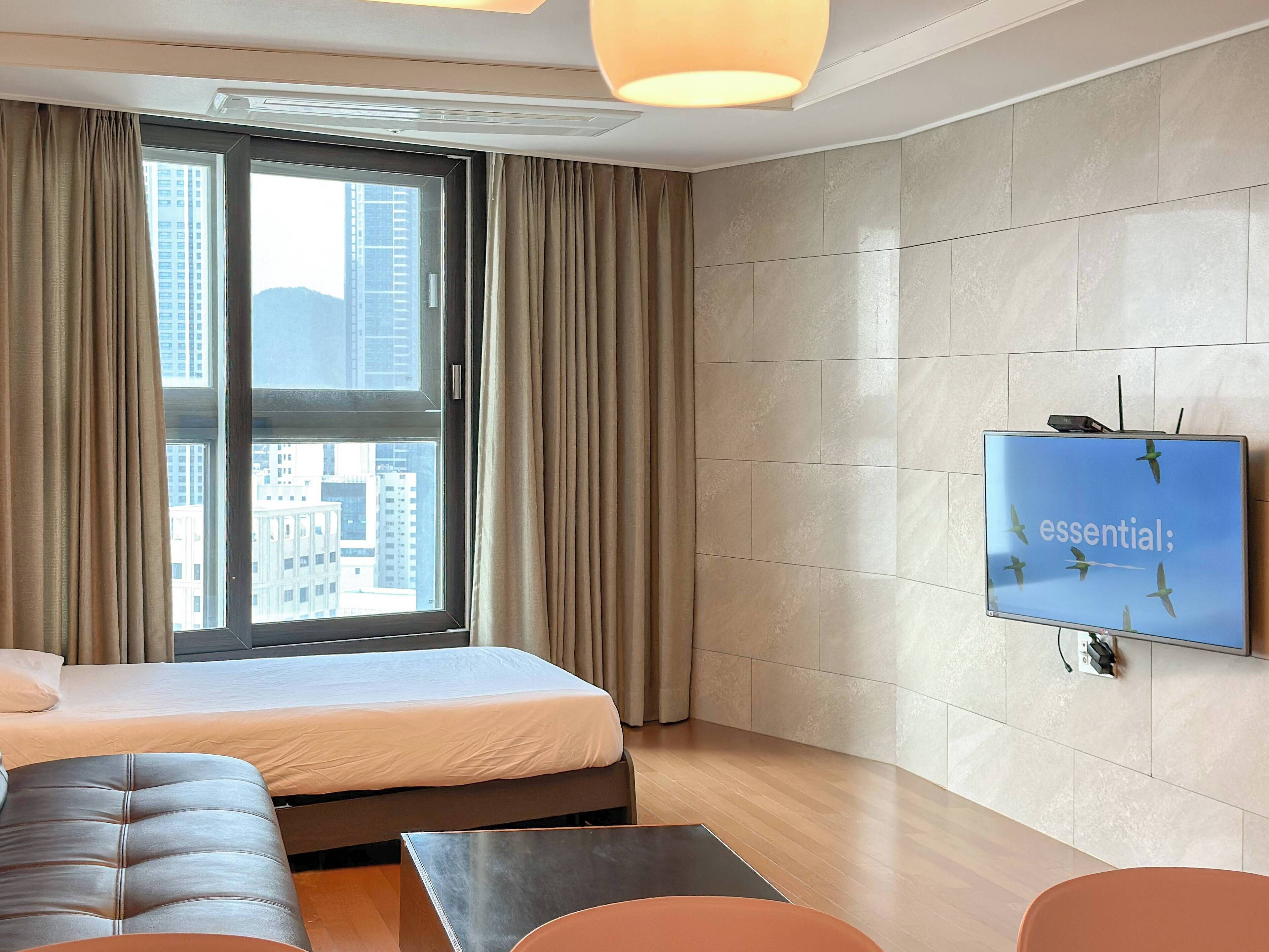 Quarto de hotel com cama, sofá, TV fixada na parede e uma janela grande com cortinas.