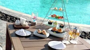 Uma mesa à beira da piscina com diversos petiscos e bebidas, incluindo um suporte para doces em vários níveis, uma jarra e copos de chá.