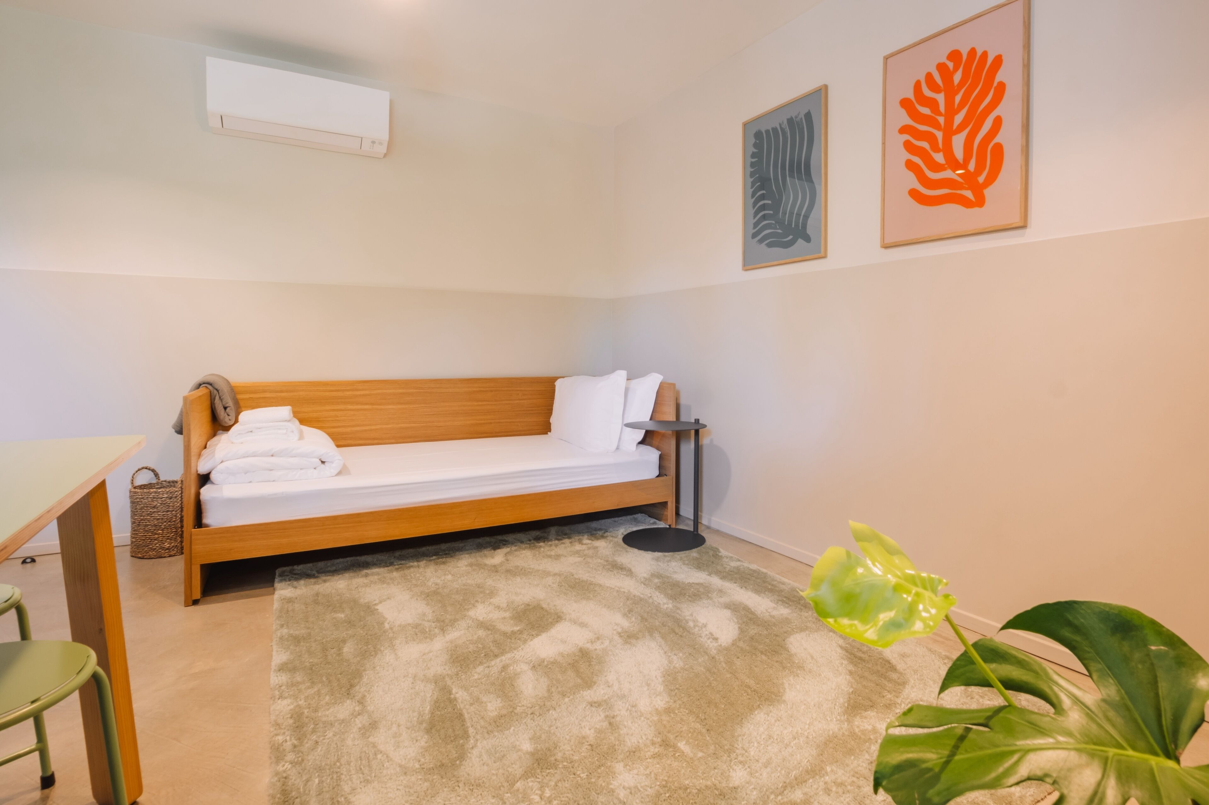 Quarto individual com uma cama de solteiro, cabeceira de madeira, uma mesinha, uma cadeira verde, um tapete cinza e uma planta.