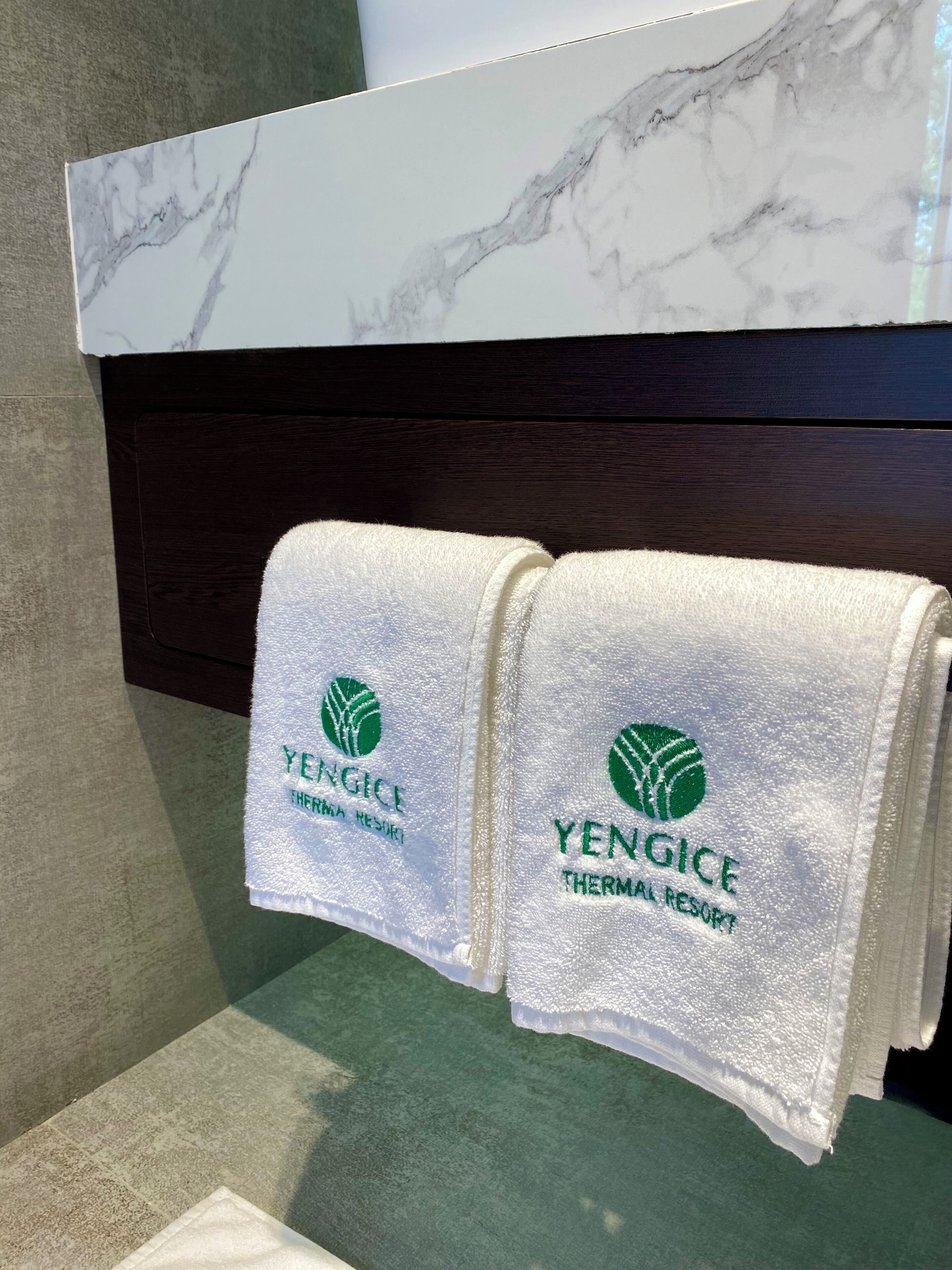 Duas toalhas com a marcação 'YENGICE THERMAL RESORT' penduradas em uma superfície de madeira escura.