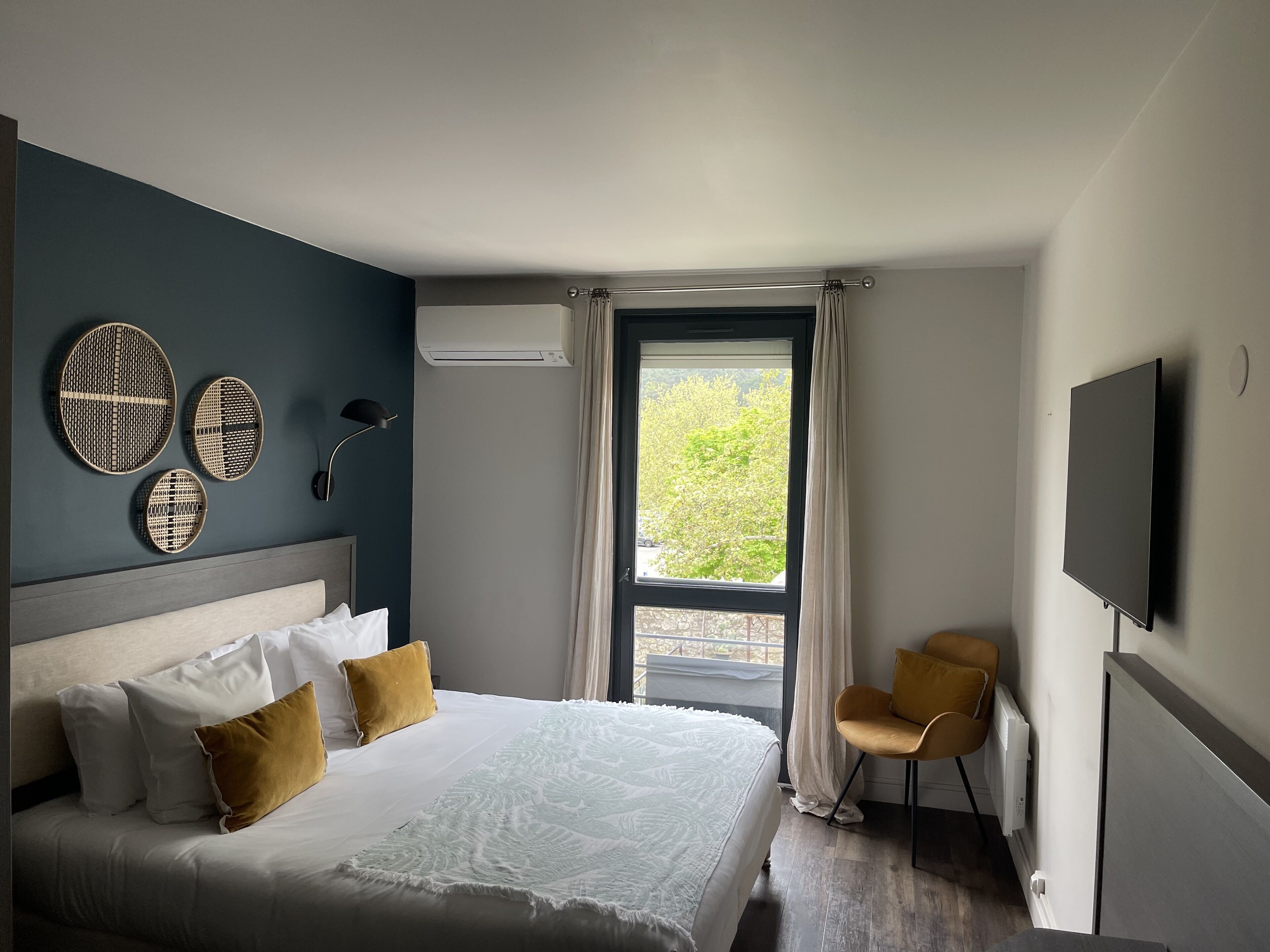 Quarto de hotel moderno com uma cama grande, televisão de tela plana, janela com cortinas e vista para o verde lá fora.