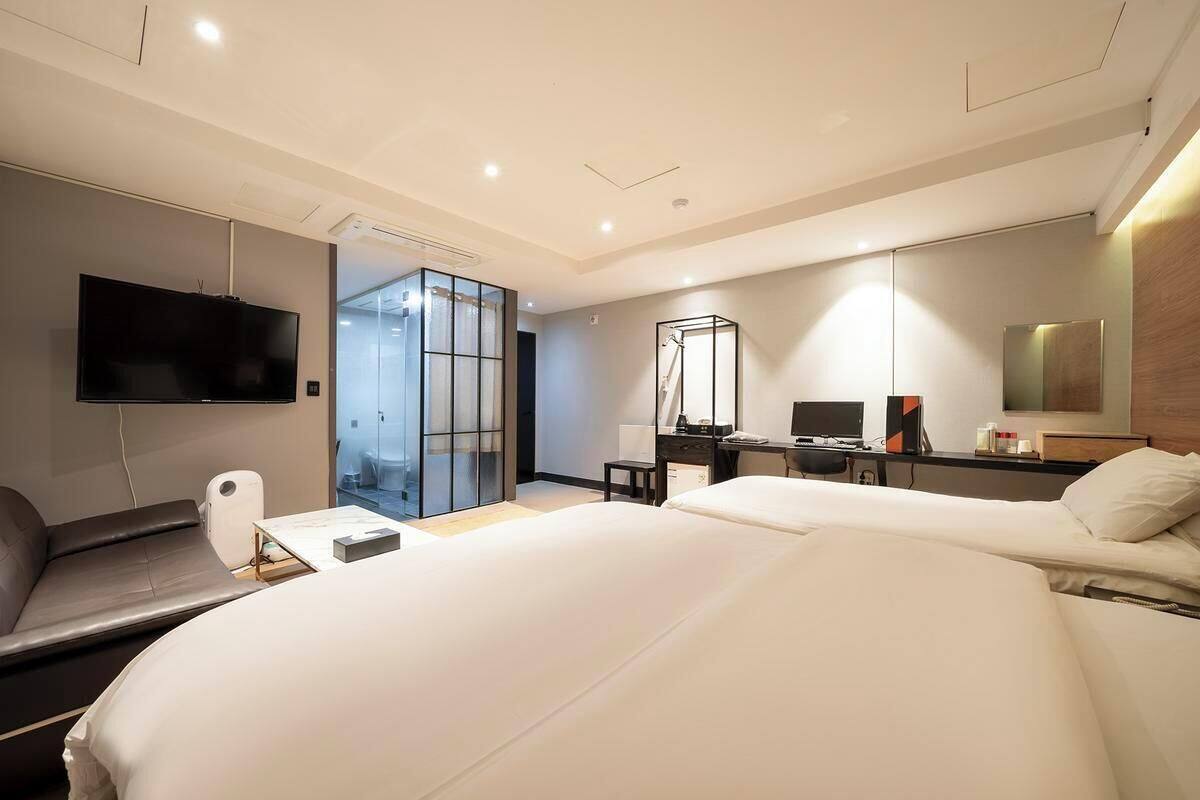 Quarto de hotel com cama, escrivaninha, cadeira, televisão e uma janela grande.