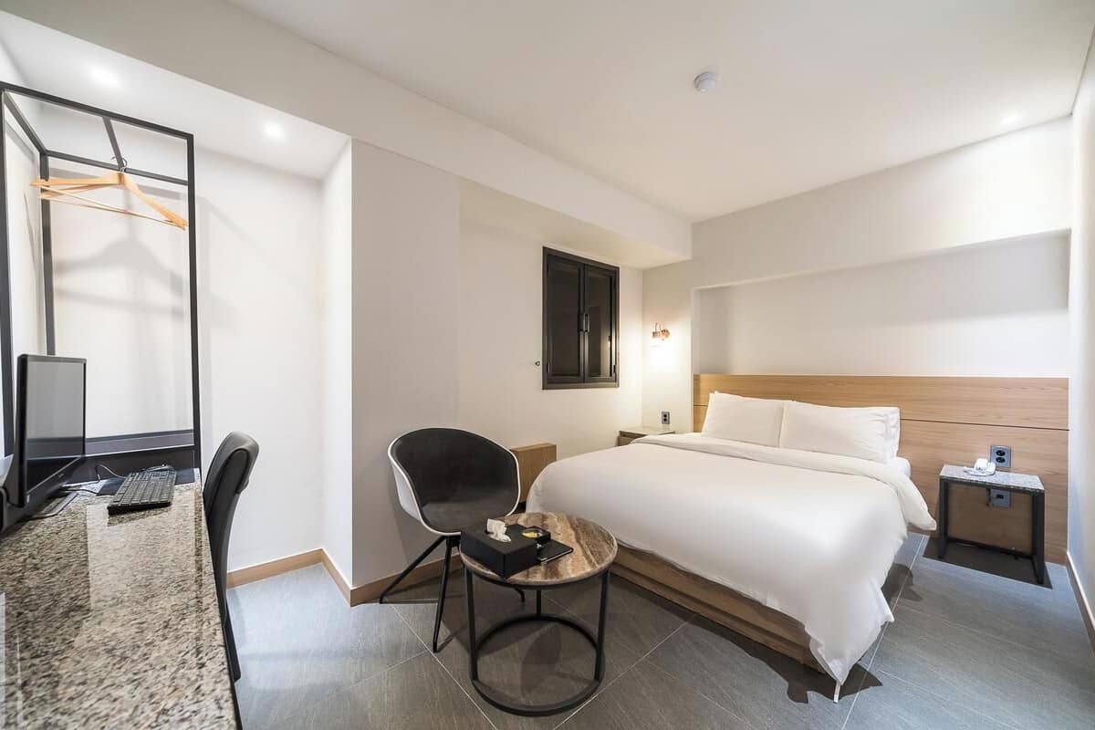 Quarto de hotel com cama, escrivaninha, cadeira, mesinha, televisão e cabeceira de madeira.