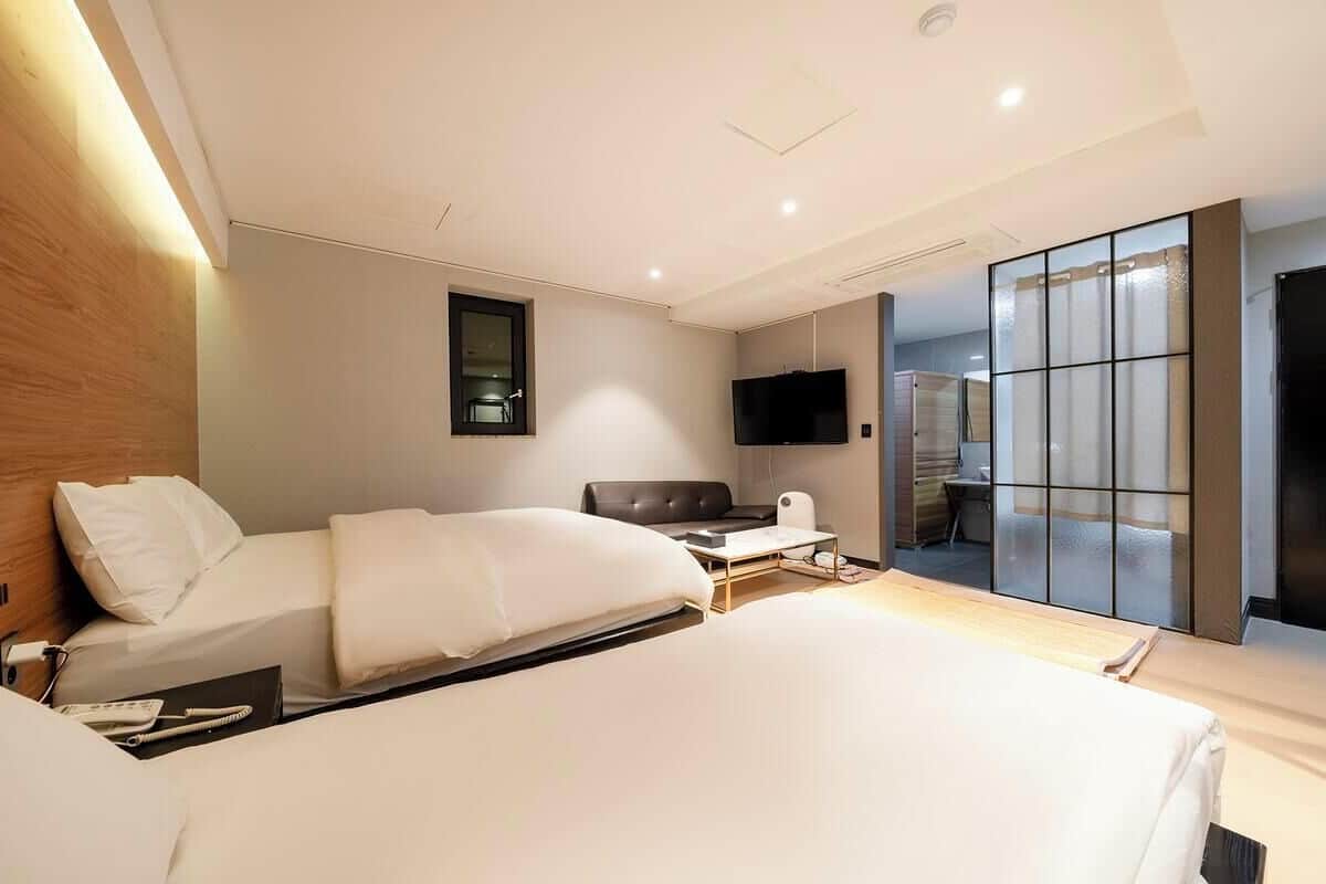 Quarto de hotel com duas camas, área de estar, televisão e uma janela grande.