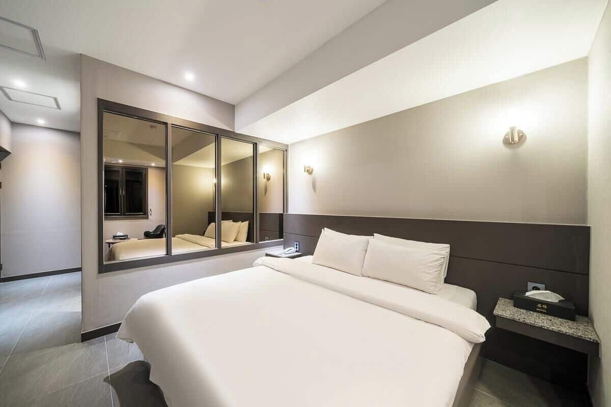 Quarto de hotel moderno com uma cama grande, uma mesa de cabeceira, um espelho e uma luminária fixada na parede.