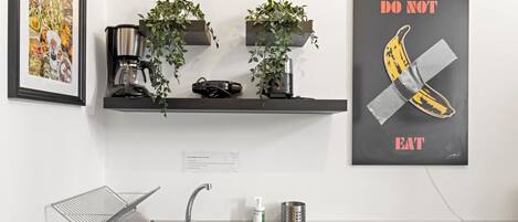 Uma cozinha com bancada de madeira, pia de aço inoxidável e máquina de café. Existem duas prateleiras sobre a pia com plantas e uma moldura com foto. Um cartaz na parede diz "NÃO COMA".