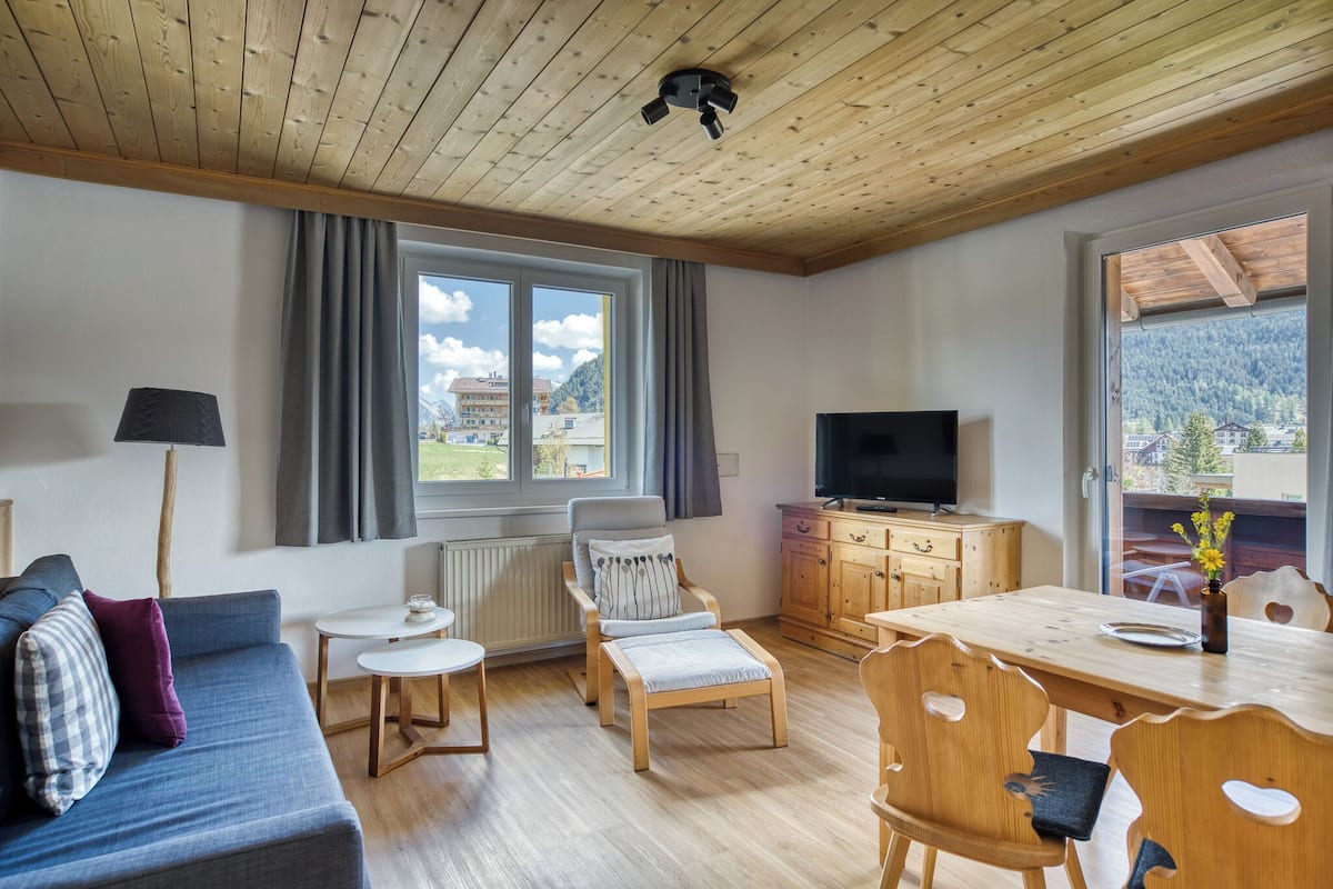 Uma sala de estar moderna com teto de madeira, um sofá, uma TV, uma mesa de jantar e vista para montanhas através de grandes janelas.