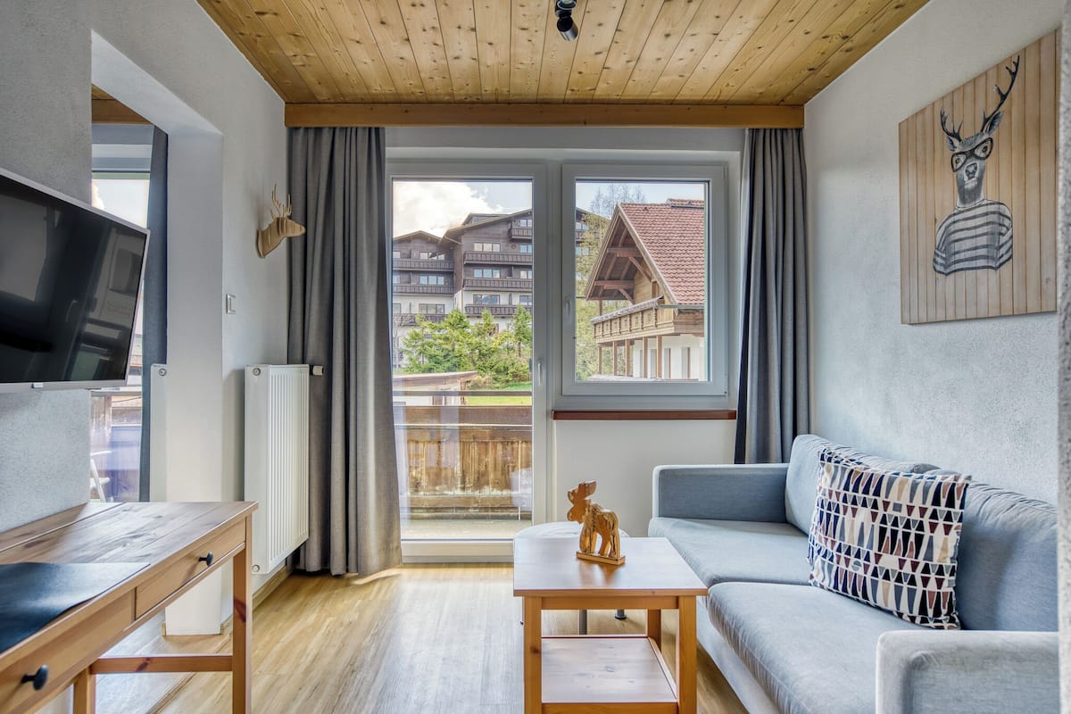 Uma sala de estar moderna com teto de madeira, uma TV, um sofá, uma mesa de centro e vista para um edifício através de uma porta de vidro.