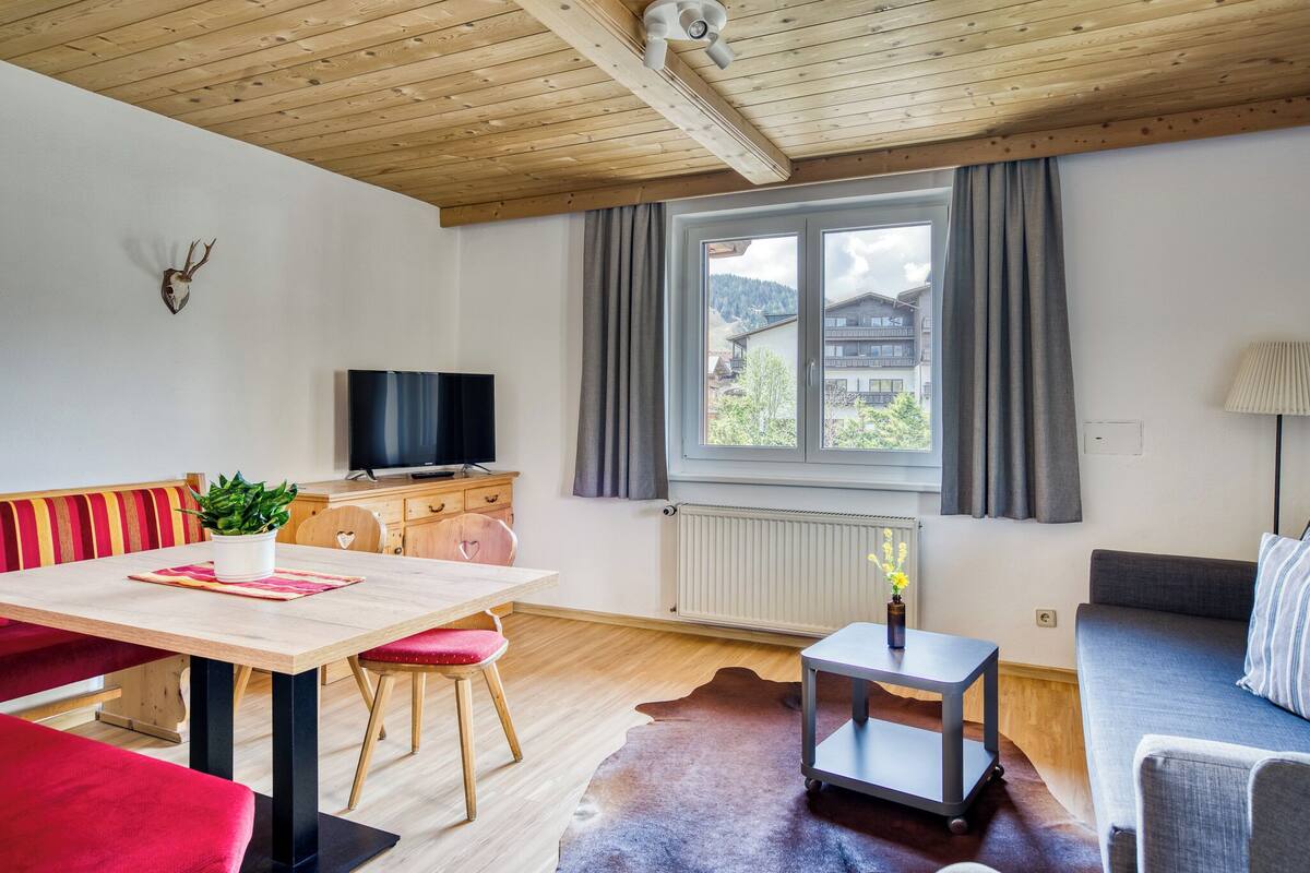 Uma sala de estar moderna com teto de madeira, um chifre de veado fixado na parede, uma televisão, uma mesa de jantar com cadeiras vermelhas, um sofá e vista para uma montanha através de uma janela.