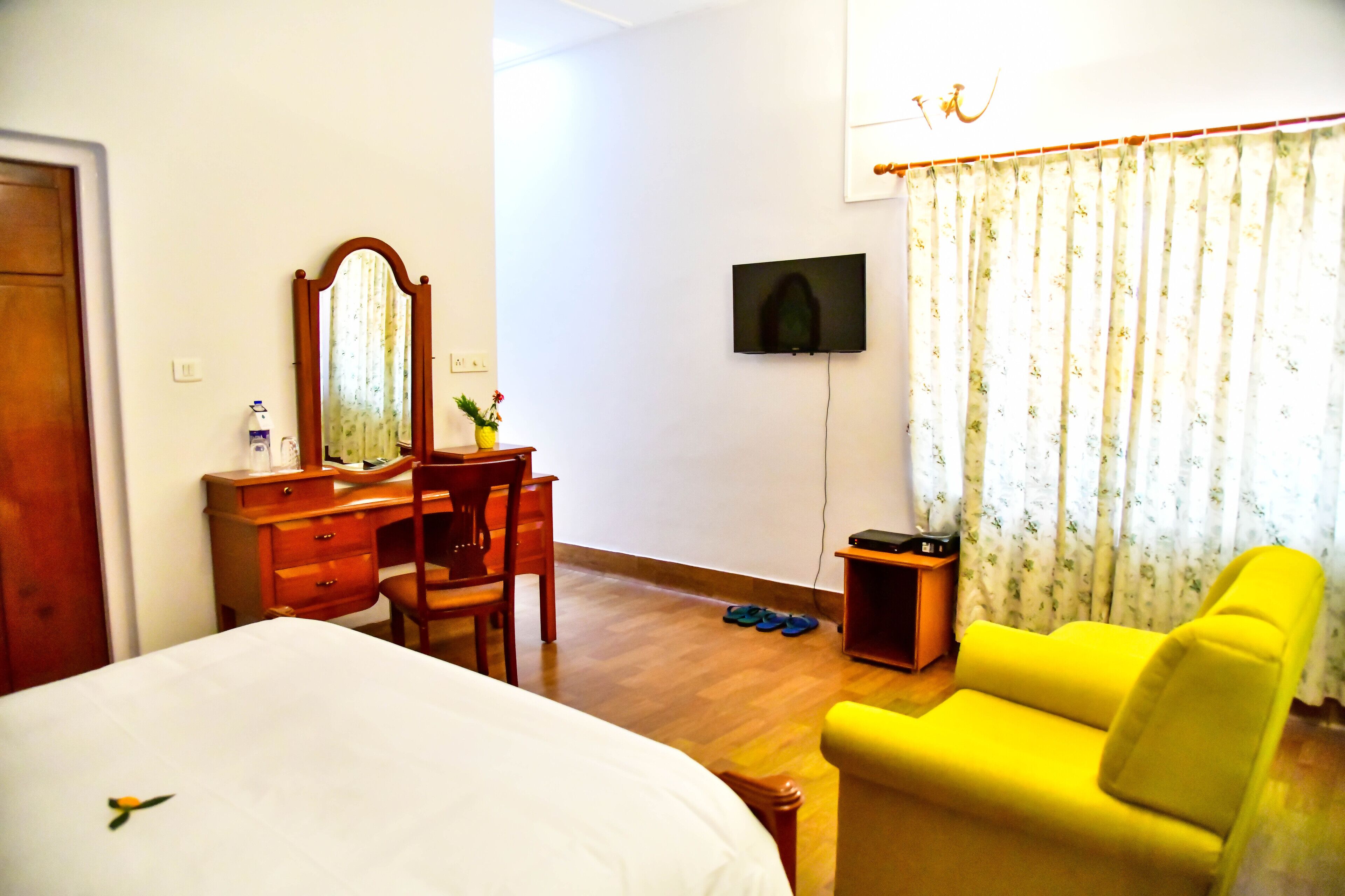 Quarto de hotel com cama, cômoda de madeira, cadeira, televisão e janela com cortinas.