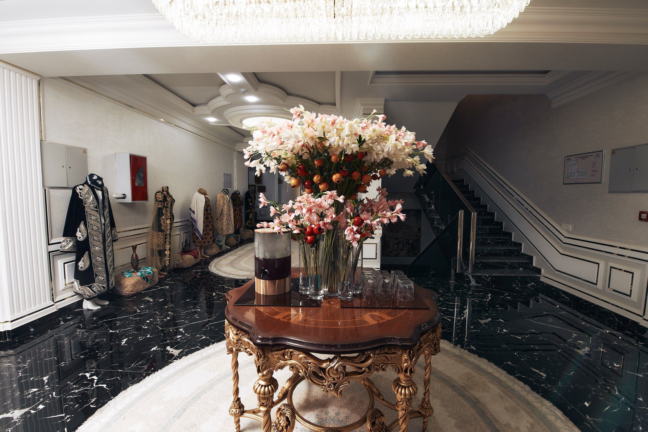 Um corredor com piso de mármore, um lustre imponente, um aparador decorativo com um vaso de flores e uma escadaria.