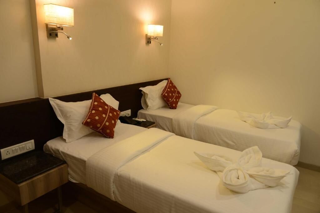 Quarto de hotel com duas camas de solteiro, cada uma com roupa de cama branca e um travesseiro com bolinhas vermelhas. Há luminárias fixadas na parede sobre cada cama.