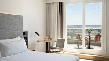 Quarto de hotel com uma cama grande, uma mesinha redonda com duas cadeiras, uma planta em vaso e vista para a cidade através de uma porta de vidro deslizante.