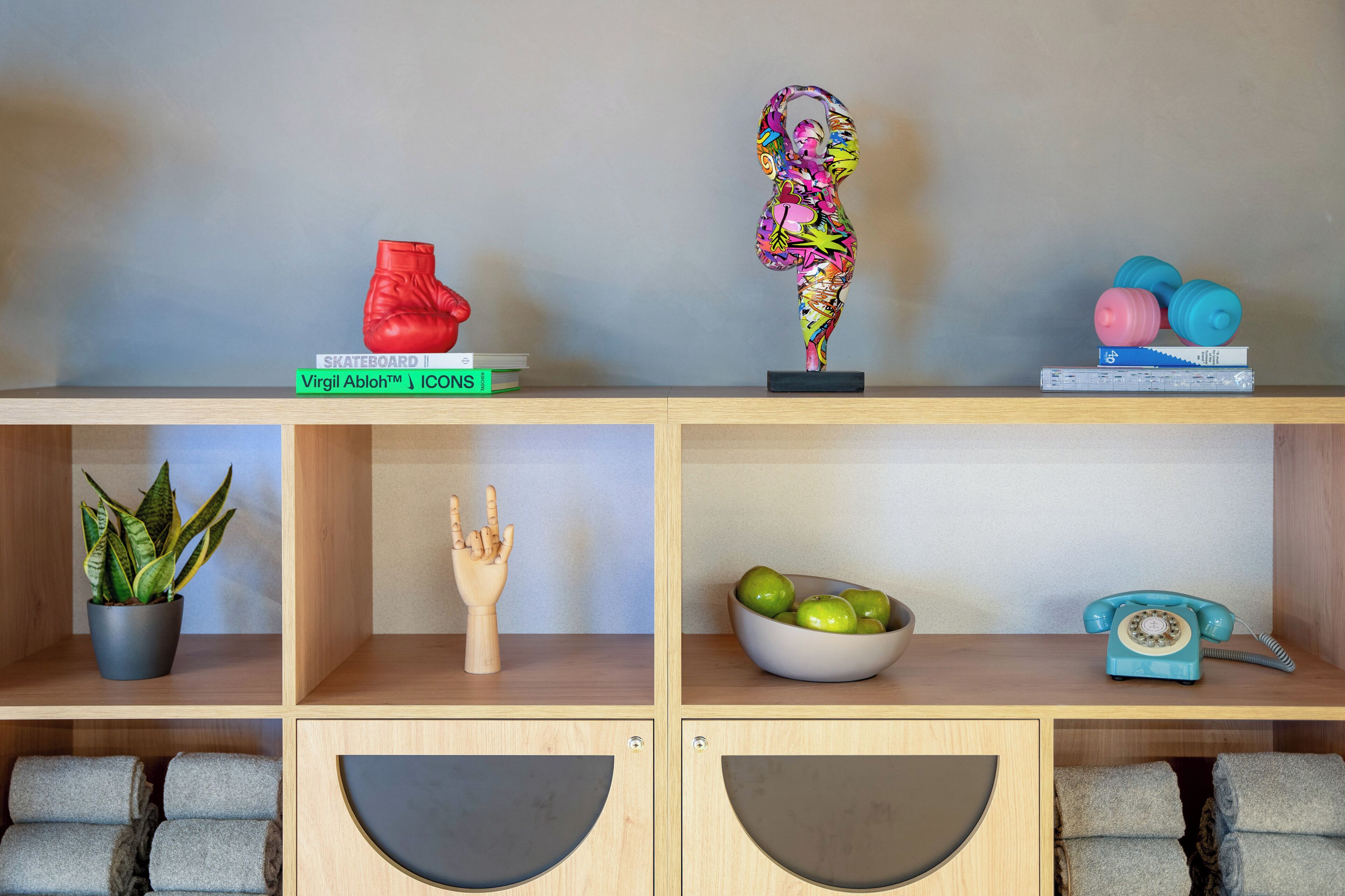 Estante de madeira com diversos objetos decorativos, incluindo uma bota vermelha, uma escultura abstrata colorida, uma tigela com maçãs verdes e um telefone azul retrô.