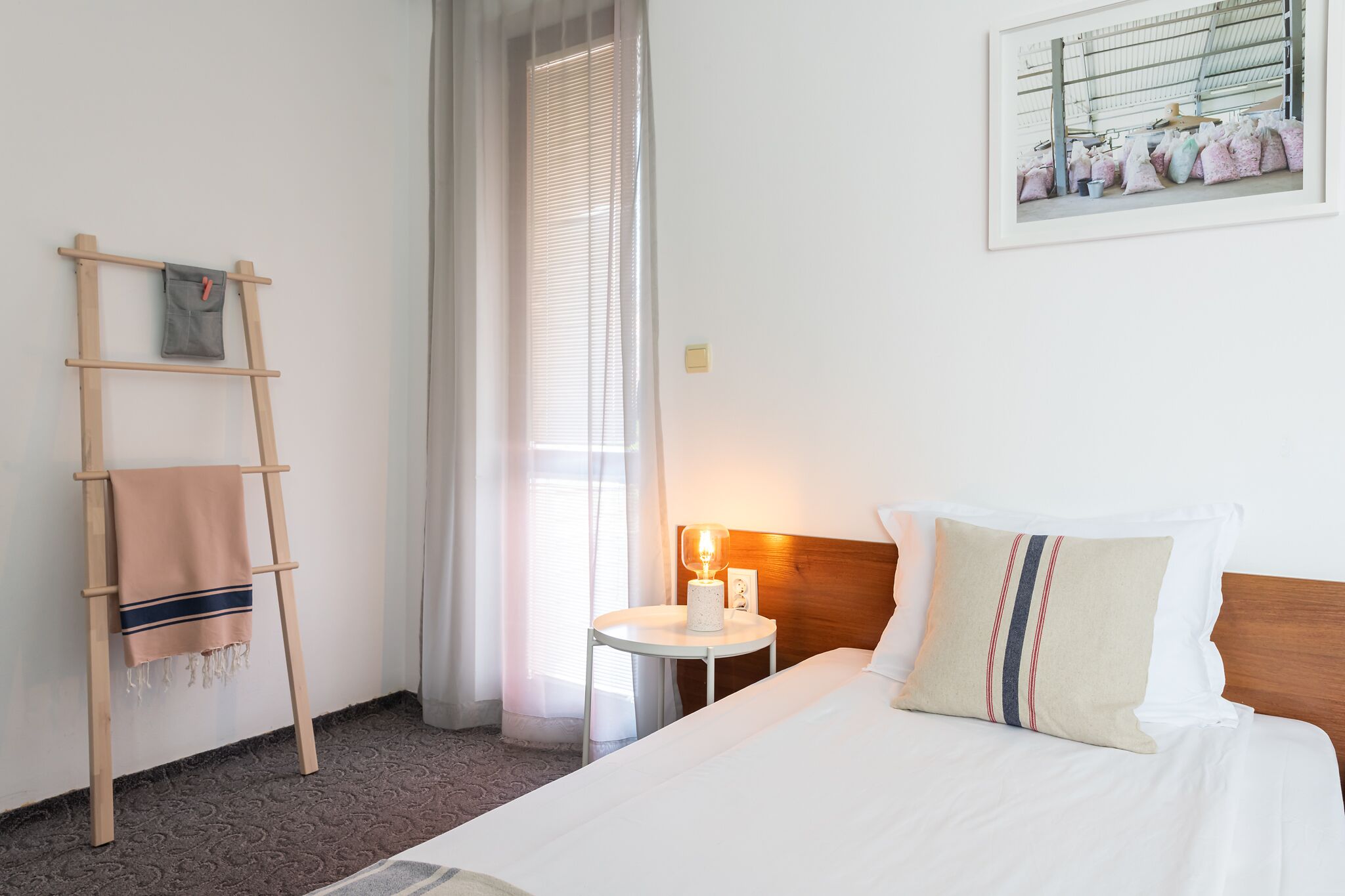 Quarto de hotel com cama, mesa de cabeceira com luminária, escada de madeira e janela com cortinas.