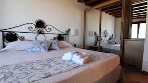 Uma cama bem arrumada com colcha estampada e travesseiros, uma mesa de cabeceira com abajur, e um espelho grande refletindo o quarto.